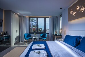 Quarto de hotel moderno com uma cama grande, duas cadeiras azuis, uma mesa de trabalho e vista da cidade através de amplas janelas.