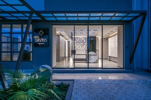 Entrada moderna de hotel com fachada de vidro, uma placa com o texto "LAVRIS City Suites" e um padrão geométrico de azulejos no chão.