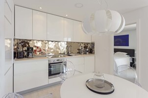 Uma cozinha moderna com armários brancos, forno de aço inoxidável e cafeteira. Há uma mesa de jantar com tampa de cúpula de vidro e um tabuleiro.