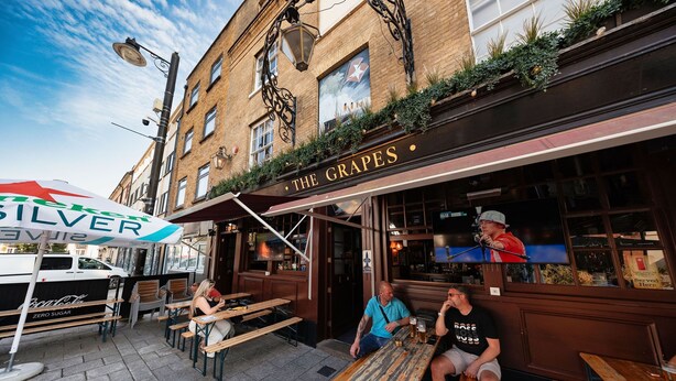 Uma área de estar ao ar livre com mesas e bancos de madeira em um restaurante chamado "The Grapes".