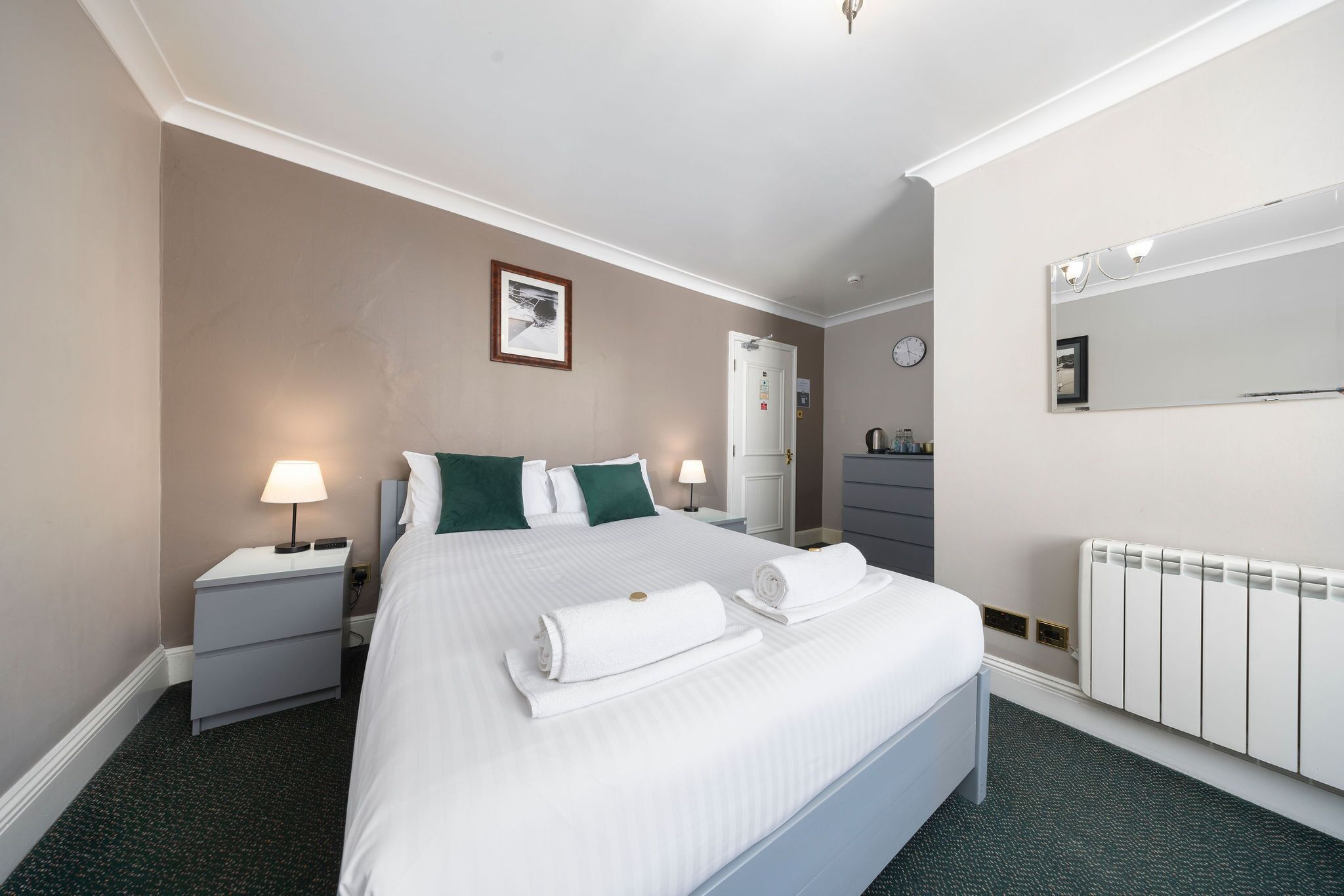 Quarto de hotel com cama bem arrumada, mesa de cabeceira com abajur, cómoda, espelho e radiador.