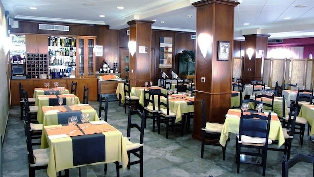 Restaurante com várias mesas e cadeiras, pilares de madeira e um bar com prateleiras cheias de garrafas.