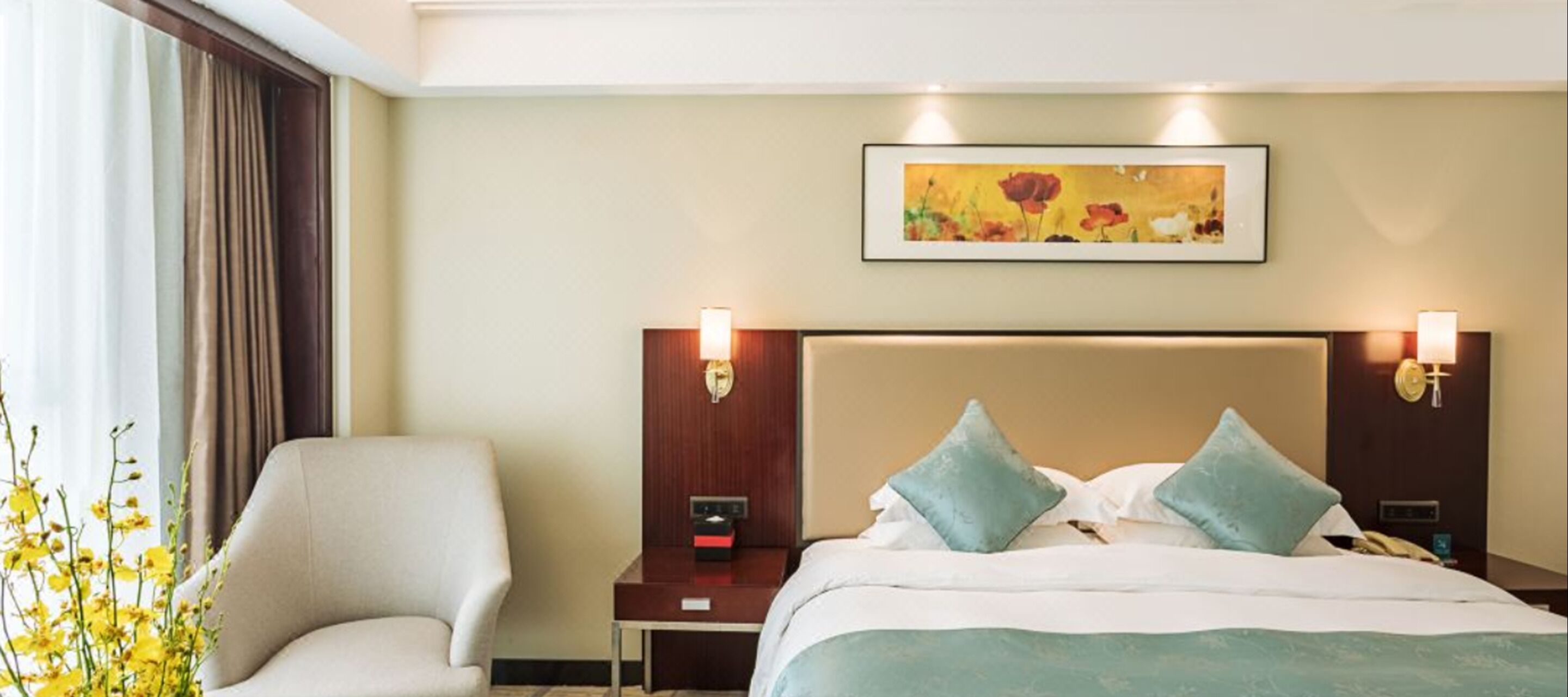 Quarto de hotel com uma cama, duas luminárias de cabeceira, uma mesa de apoio, uma cadeira e um quadro na parede.