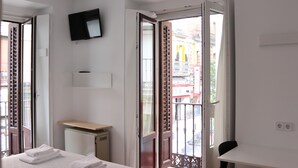Quarto de hotel com cama, escrivaninha, televisão na parede e varanda com vista para a cidade.