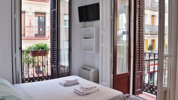 Quarto de hotel com cama, televisão na parede, janela com vista para a rua e varanda com planta.