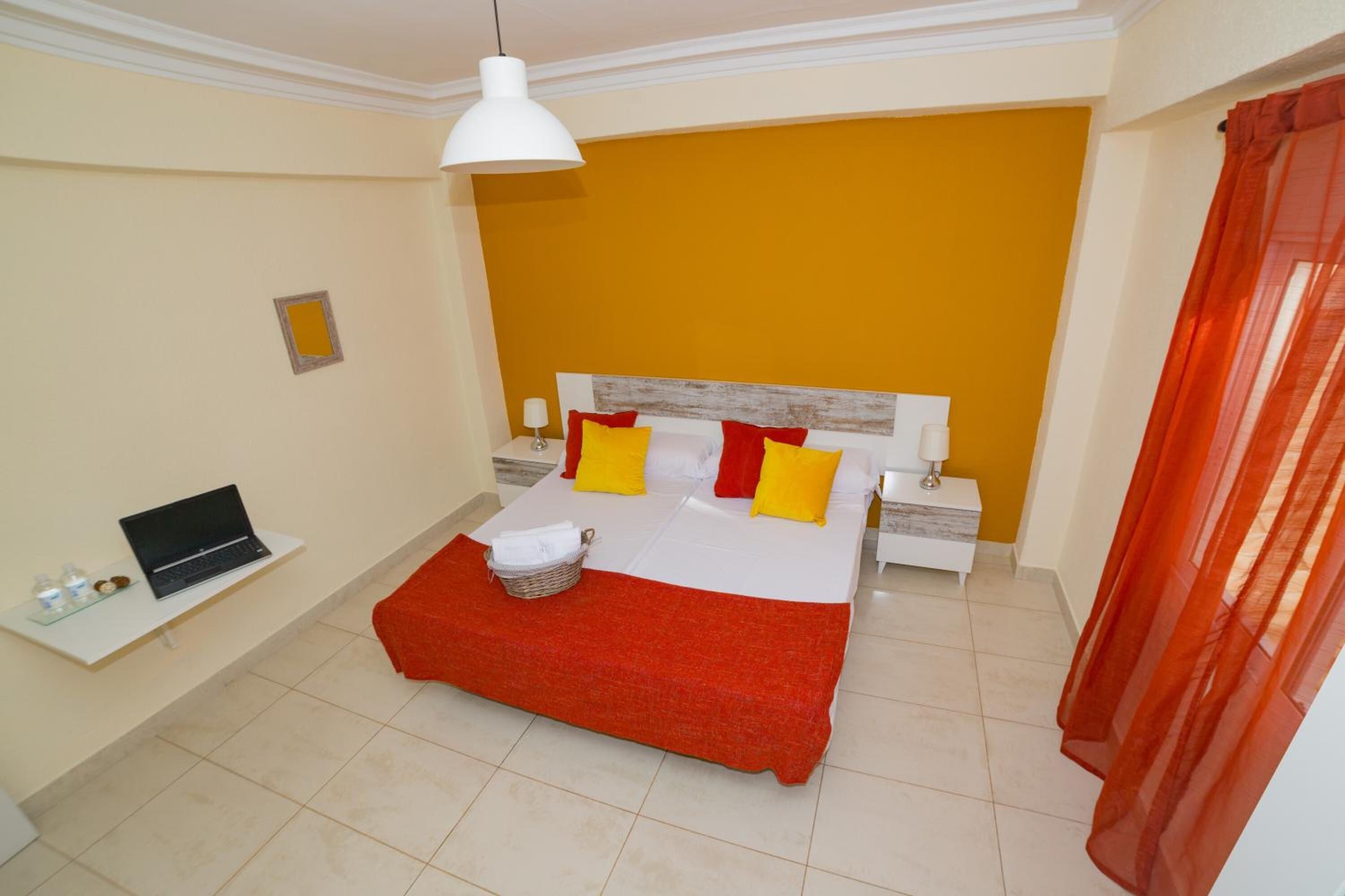 Quarto de hotel com cama, laptop numa estante, mesa de cabeceira com abajur e cortina vermelha.