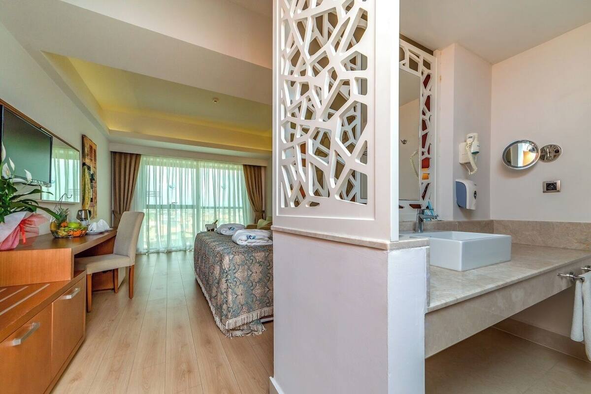 Quarto de hotel com escrivaninha de madeira, cama com colcha estampada, janela grande com cortinas, banheiro com pia e espelho, e divisória branca decorativa.