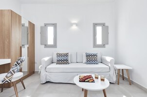 Uma sala de estar moderna com um sofá branco, duas almofadas decorativas estampadas, uma mesa de centro com petiscos e um guarda-roupa de madeira.