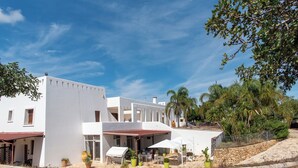 Um edifício moderno e branco com um pátio coberto, mesas ao ar livre e plantas em vasos.