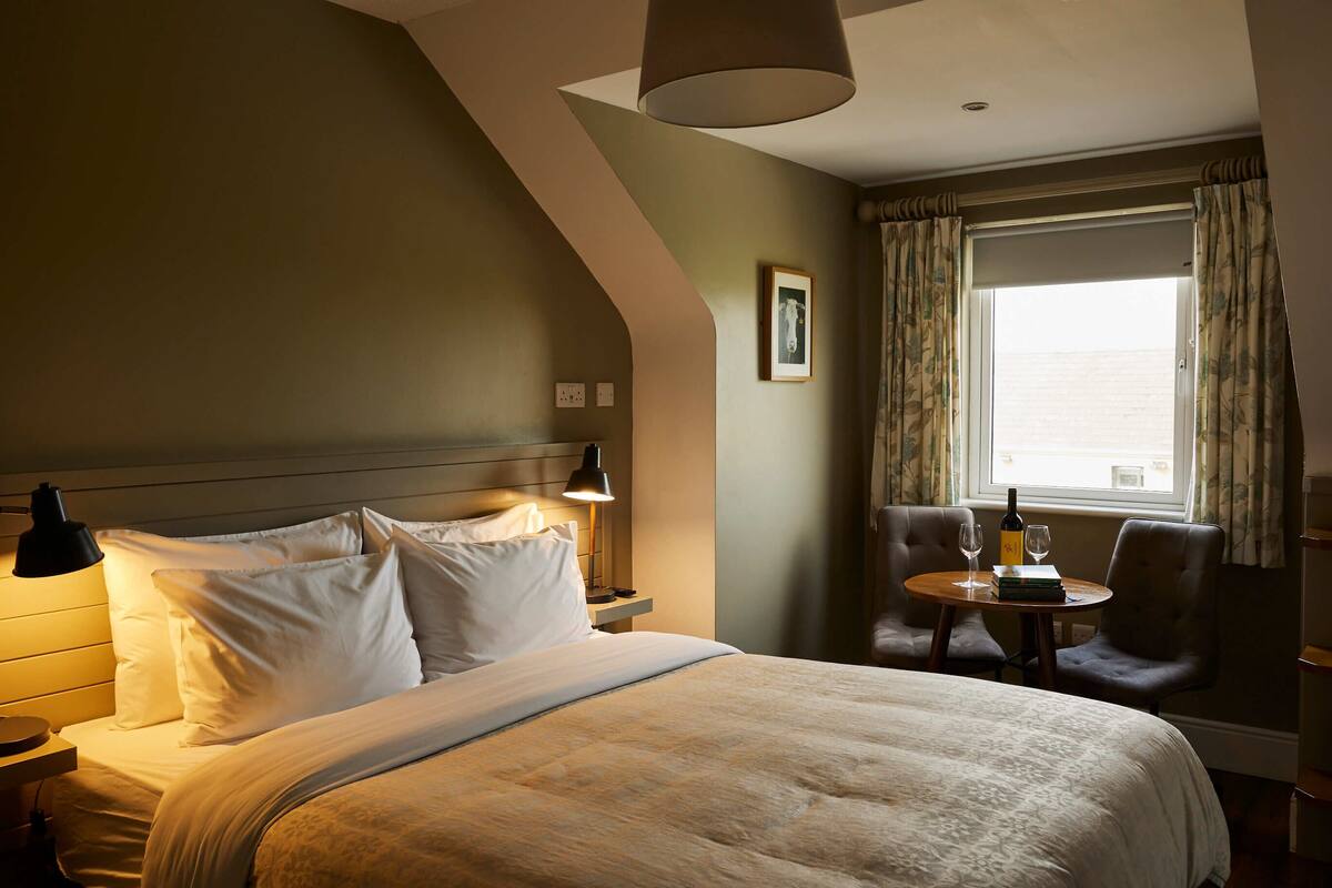 Quarto de hotel com cama, mesa de cabeceira, luminária, cadeira, pequena mesa com garrafa e copo, e janela com cortinas.