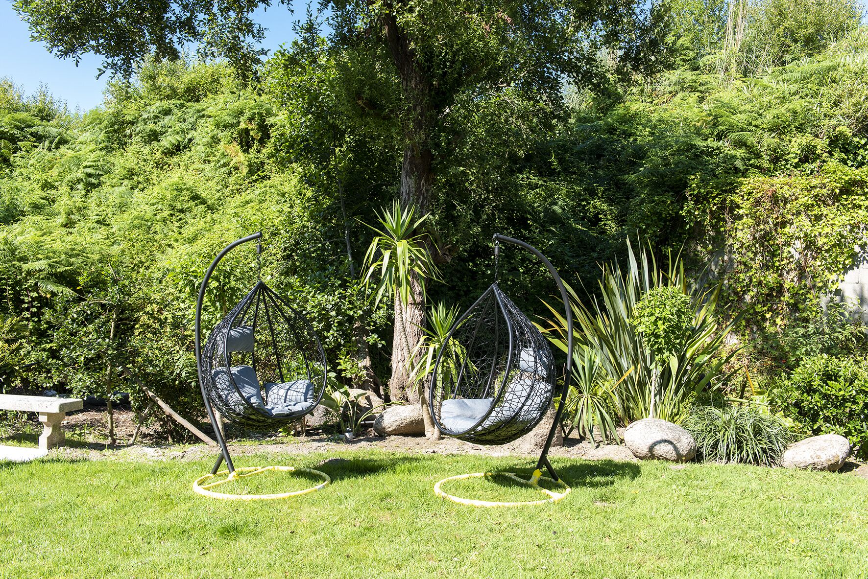 Um jardim com duas cadeiras suspensas, um banco de pedra e diversas plantas.