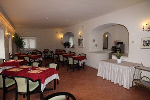 Uma sala ampla com paredes de tijolo e chão de azulejos, apresentando uma área de refeições com várias mesas cobertas por toalhas quadriculadas vermelhas e brancas.