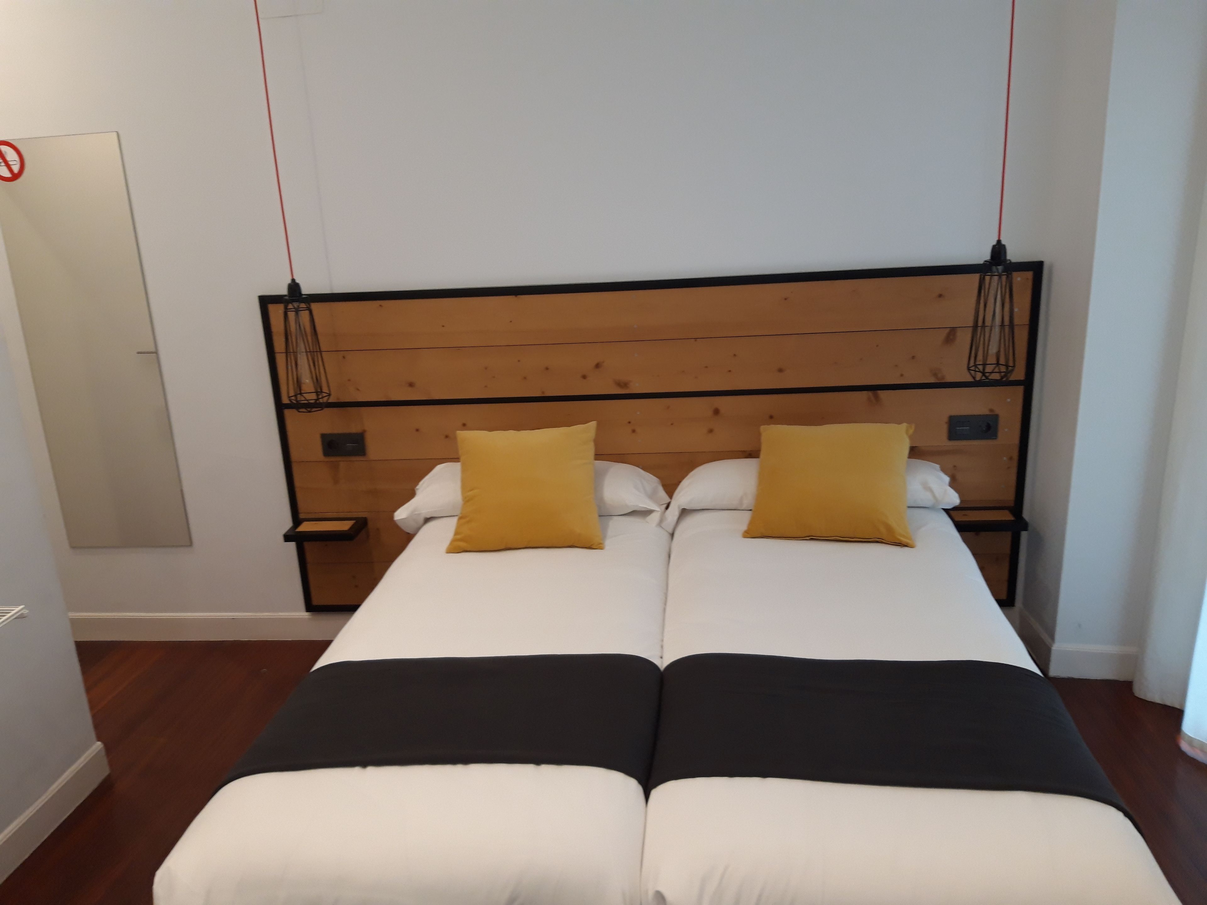 Uma cama de casal com duas almofadas, cabeceira de madeira, mesas de cabeceira e um candeeiro suspenso.