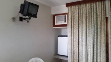 Um quarto pequeno com televisão na parede, um refrigerador e uma janela com cortinas.