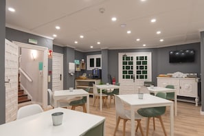Uma área de refeições moderna com mesas brancas e cadeiras verdes, uma cozinha com armários brancos e uma escada que leva a outro nível.