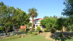 Um jardim bem cuidado com diversas árvores, incluindo uma palmeira, e um edifício com telhado vermelho ao fundo.