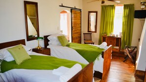 Quarto com duas camas de madeira, uma escrivaninha de madeira, um espelho e uma janela com cortinas verdes.