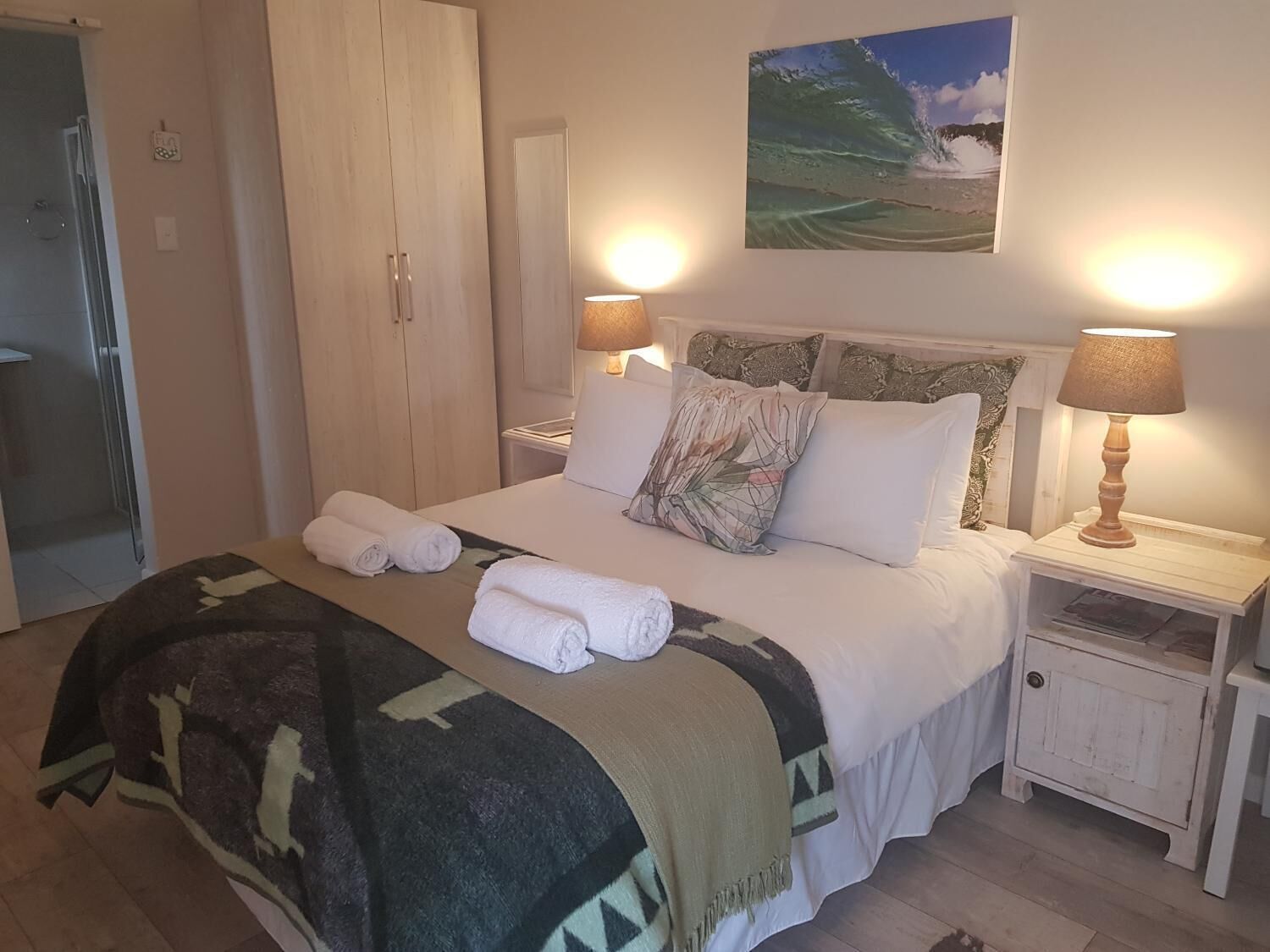 Cama bem arrumada com lençóis brancos, duas luminárias de cabeceira, um cobertor e um quadro de uma onda na parede.