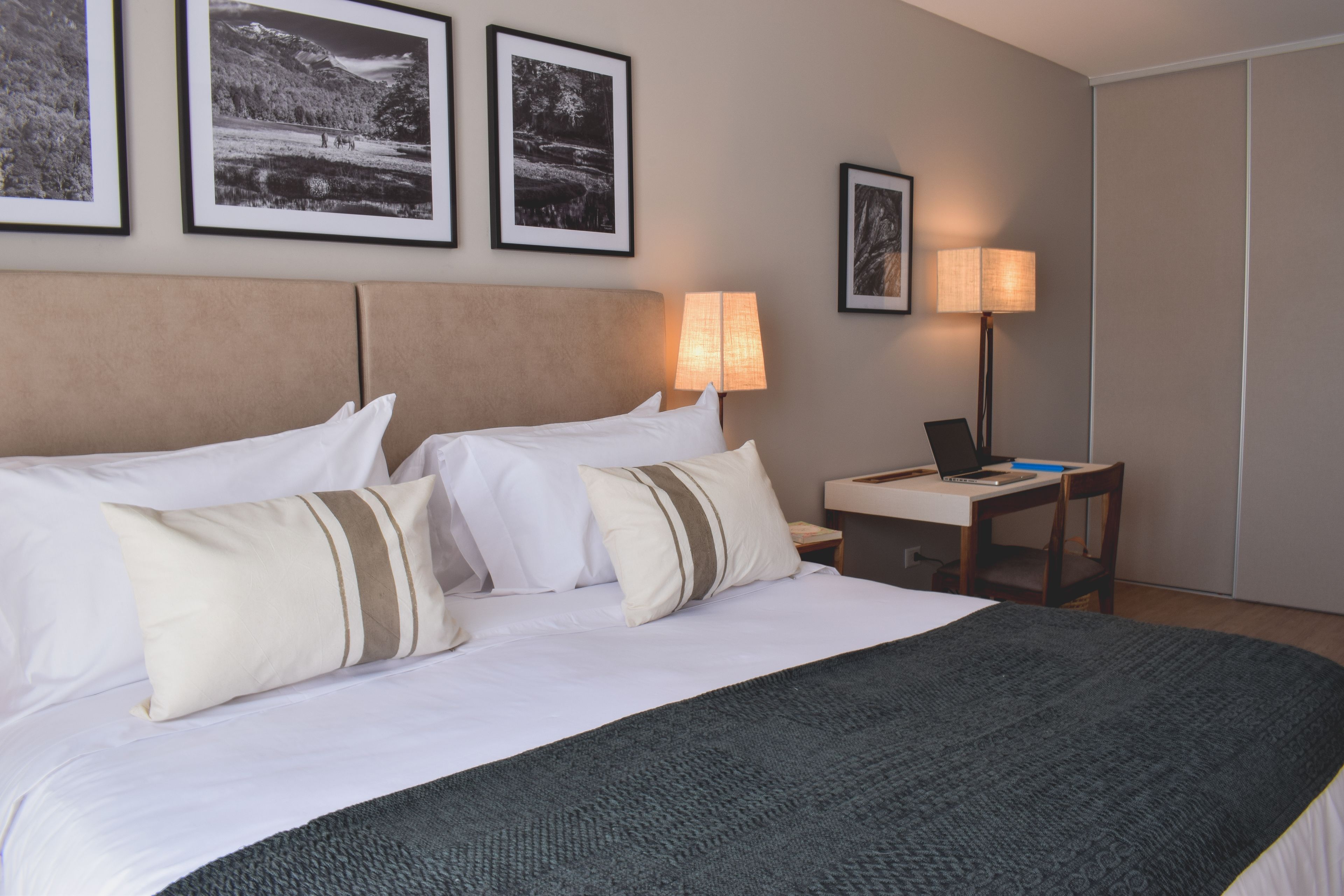 Quarto de hotel com uma cama grande, duas mesas de cabeceira, uma escrivaninha com um laptop e quadros nas paredes.