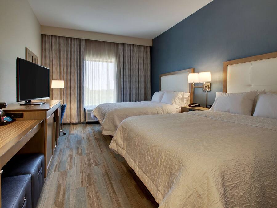 Quarto de hotel com duas camas, uma escrivaninha, televisão e janela com cortinas.