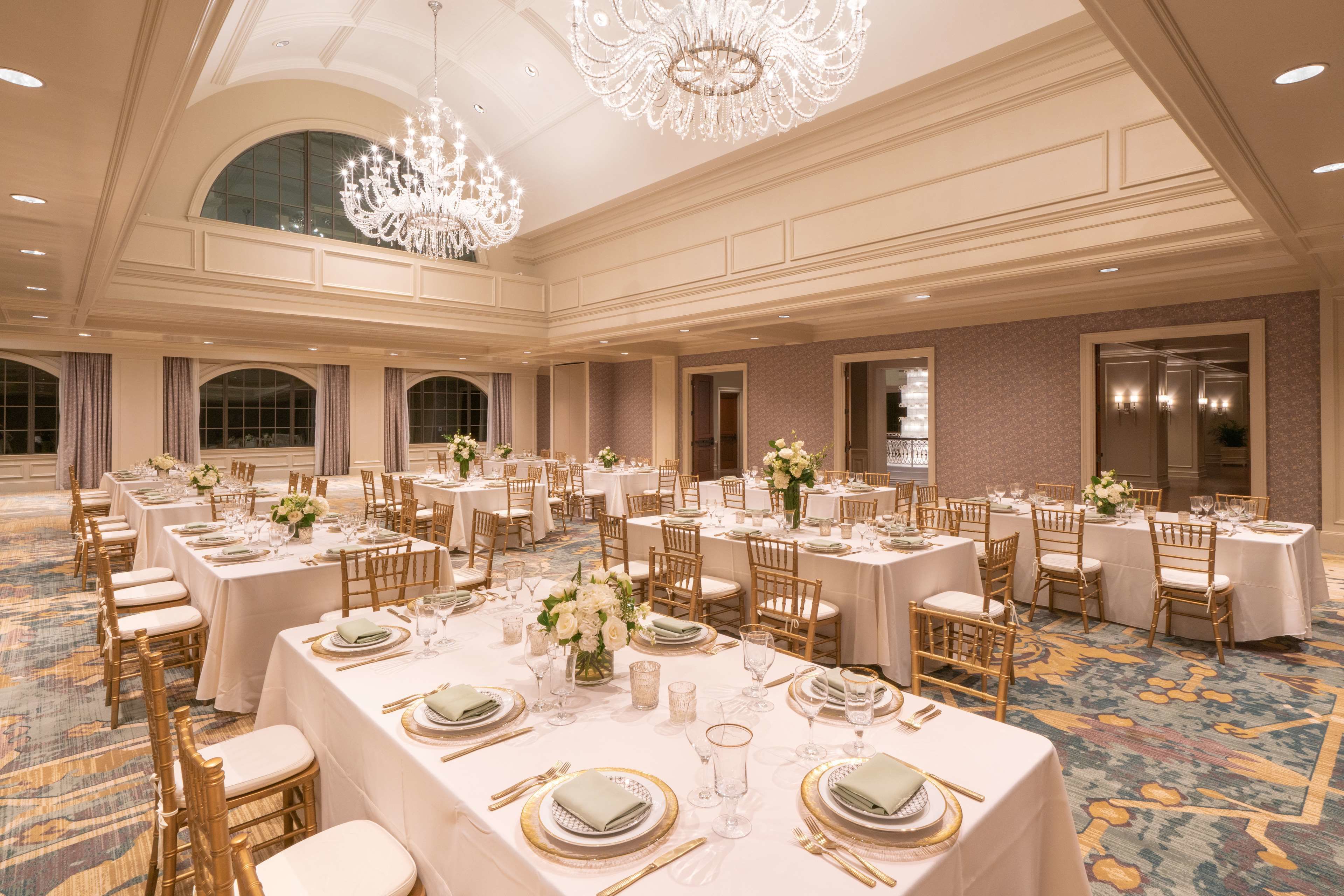 Uma sala de banquetes com várias mesas redondas, cada uma com toalhas brancas, talheres dourados e arranjos florais.