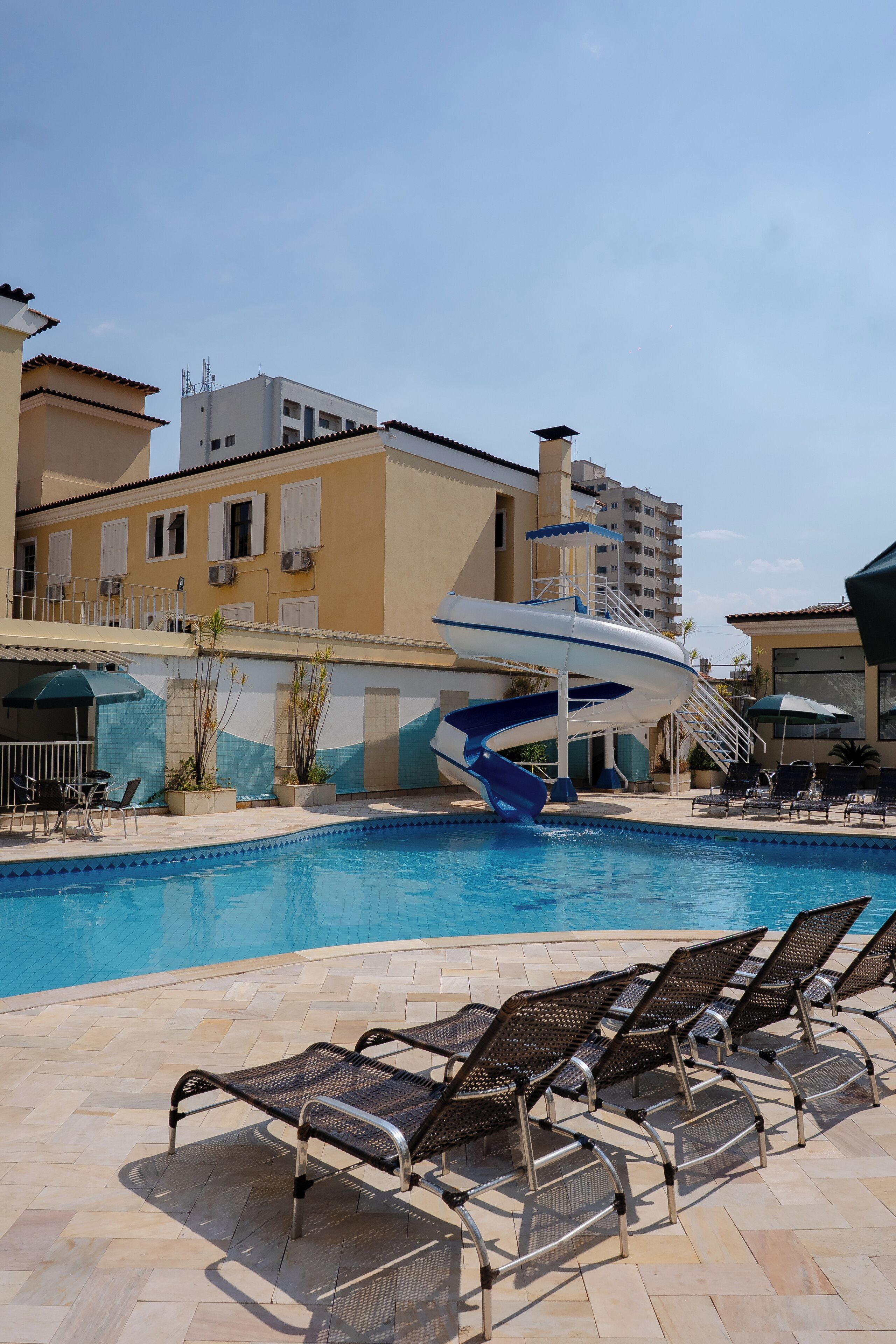 Piscina com espreguiçadeiras, tobogã e um edifício com varandas ao fundo.