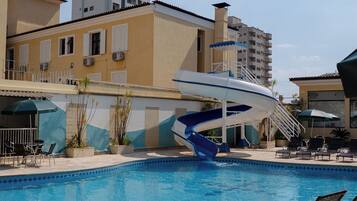 Piscina com espreguiçadeiras, tobogã e um edifício com varandas ao fundo.
