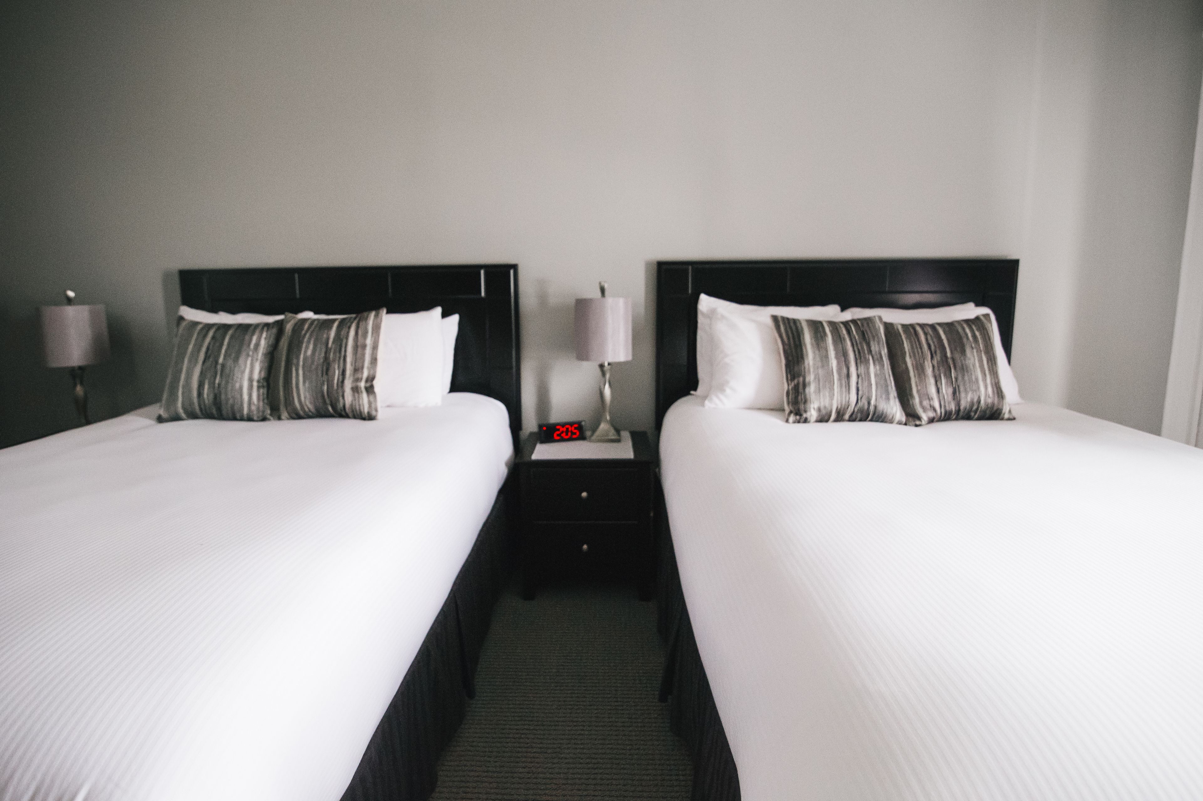 Quarto de hotel com duas camas, cada uma com roupa de cama branca e almofadas listradas, e mesinhas de cabeceira com luminárias.