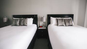 Quarto de hotel com duas camas, cada uma com roupa de cama branca e almofadas listradas, e mesinhas de cabeceira com luminárias.