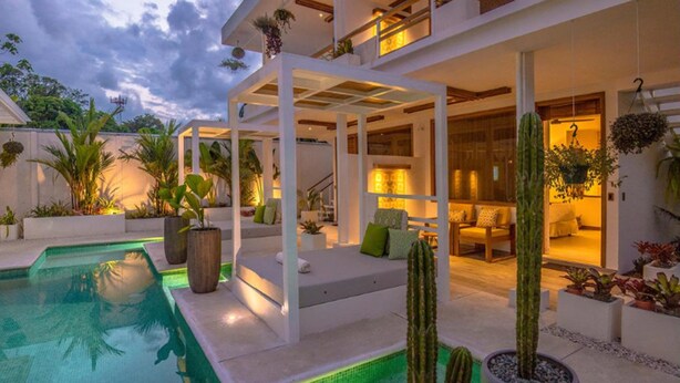 Uma área de piscina moderna ao ar livre, com espaço coberto para sentar, plantas em vasos e um espaço interno bem iluminado.