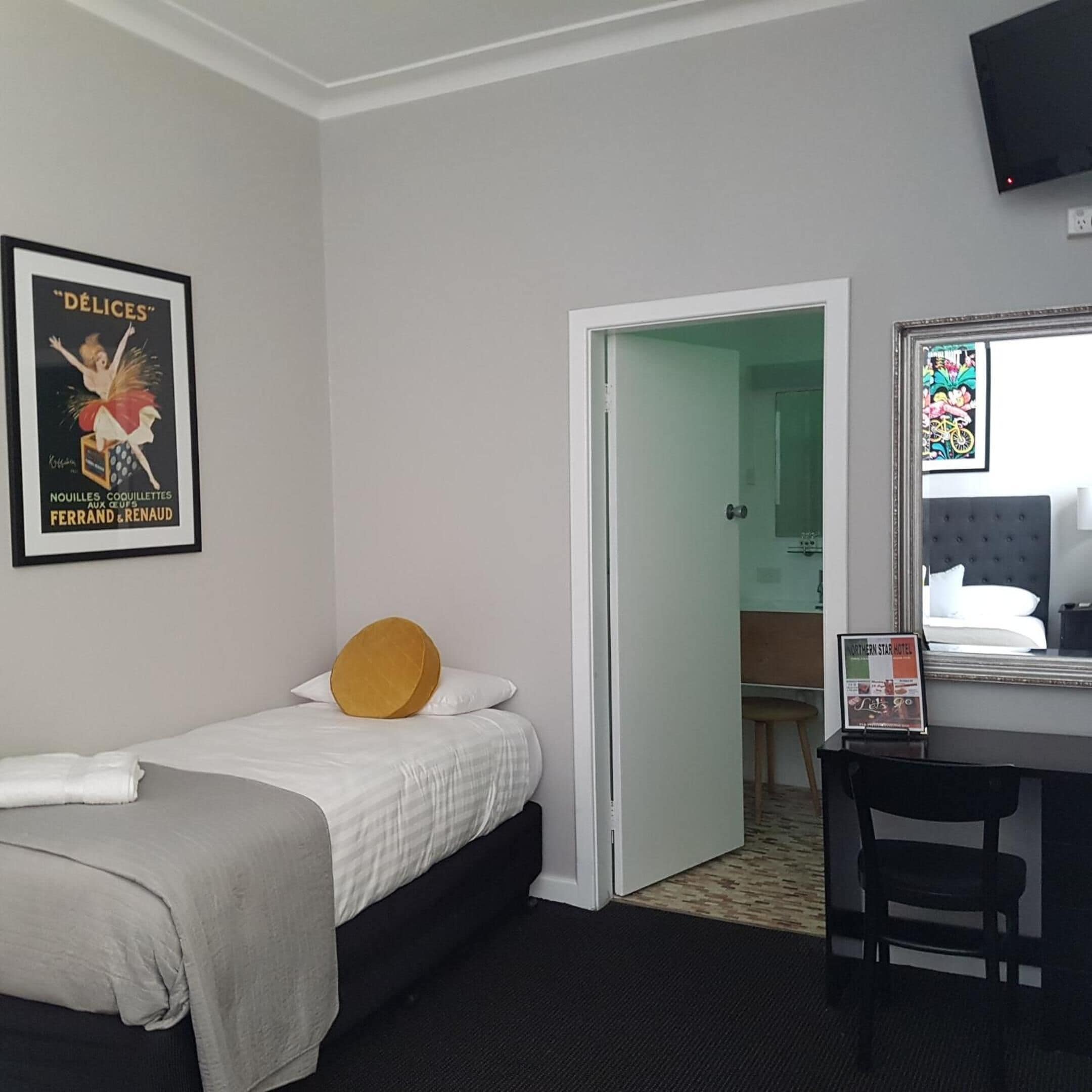 Quarto de hotel com uma cama, uma escrivaninha e uma cadeira. Há um cartaz na parede e uma porta que dá para outro quarto.