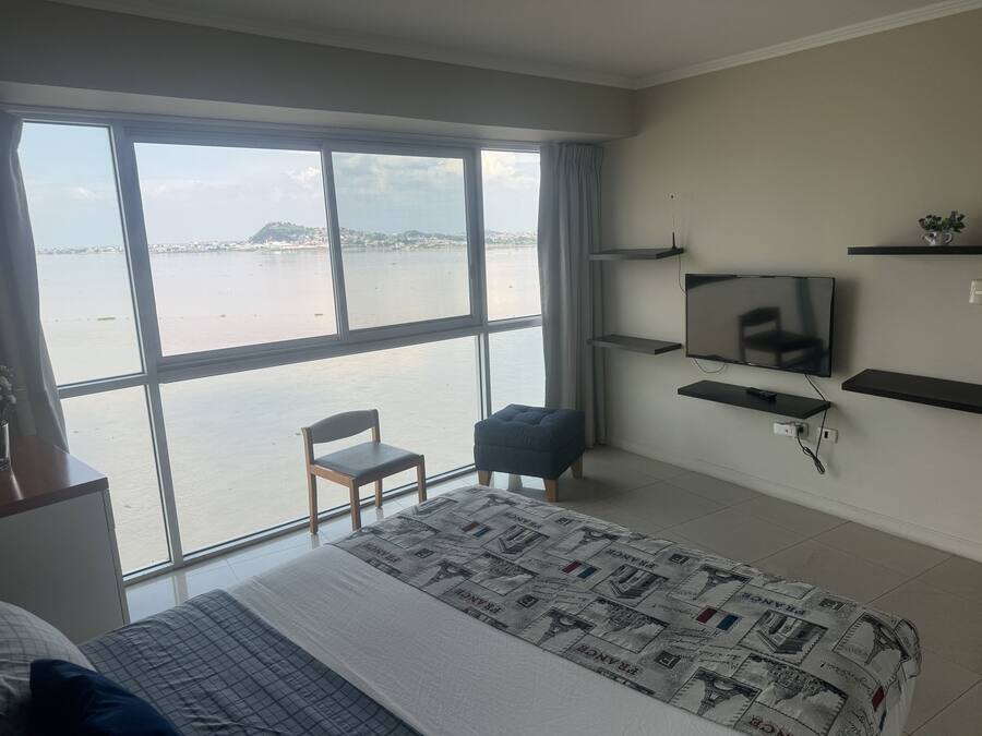 Quarto de hotel com uma grande janela que oferece vista para um corpo d'água e terra distante. Há uma cama com colcha estampada, uma cadeira e uma televisão montada na parede.