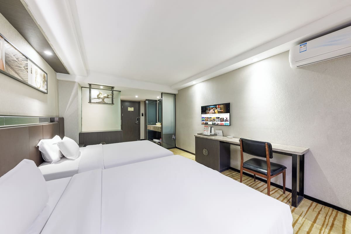 Quarto de hotel com uma cama grande, uma escrivaninha com cadeira, uma pequena cozinha compacta e um banheiro visível por uma porta aberta.