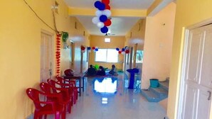 Um corredor com cadeiras de plástico vermelho alinhadas em um lado, uma mesa com cadeiras no centro e um ventilador de teto.