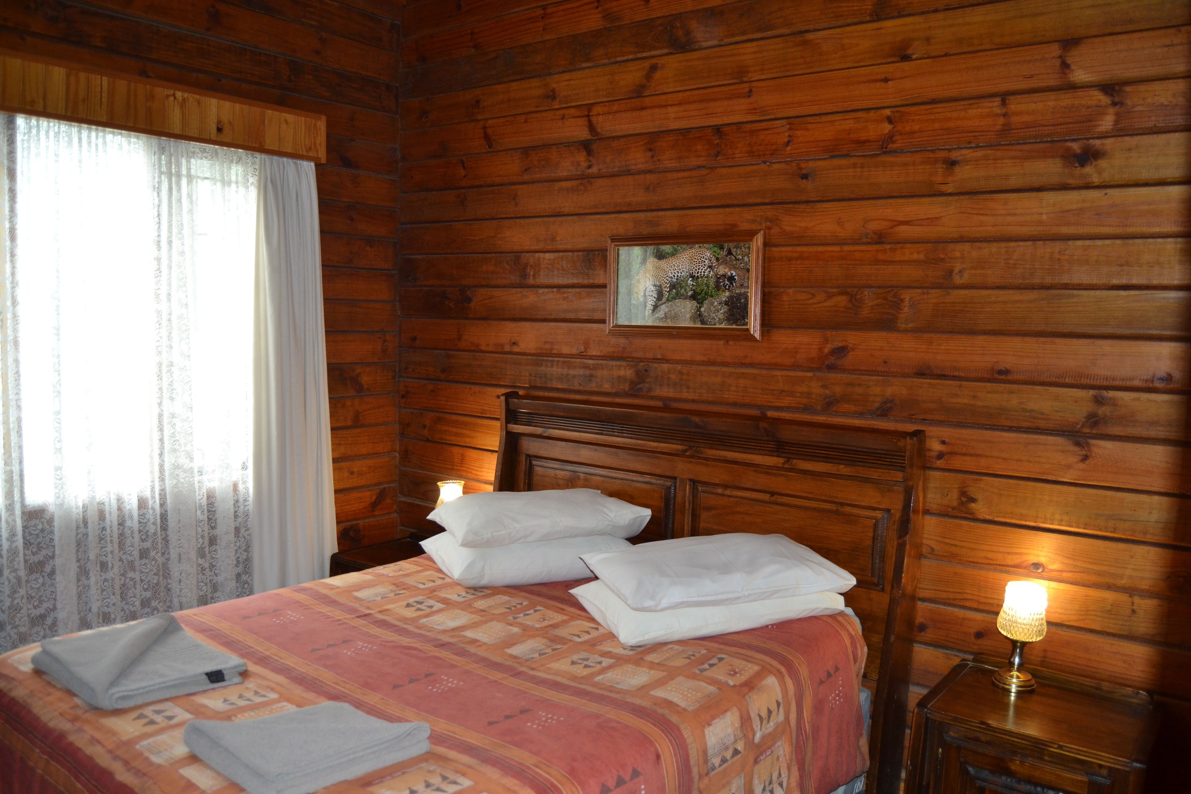 Quarto com painéis de madeira, cama, travesseiros, mesa de cabeceira com luminária e janela com cortinas.