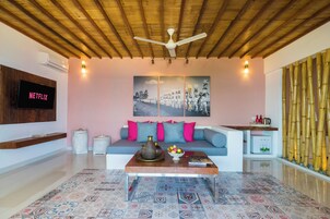 Uma sala de estar com teto de madeira, ventoinha no teto, televisão de tela plana, sofá com almofadas, mesa de centro com objetos decorativos e um tapete estampado.
