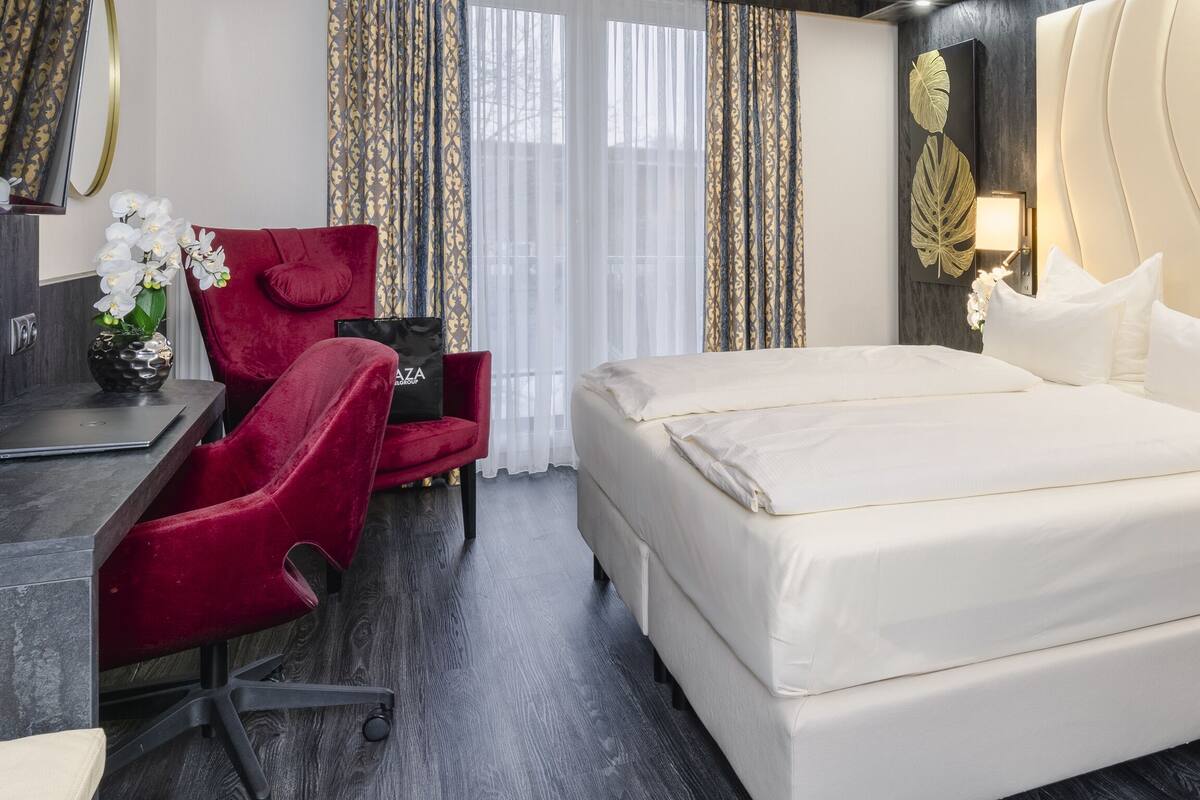 Quarto de hotel com cama, escrivaninha, cadeira vermelha e um espelho grande.