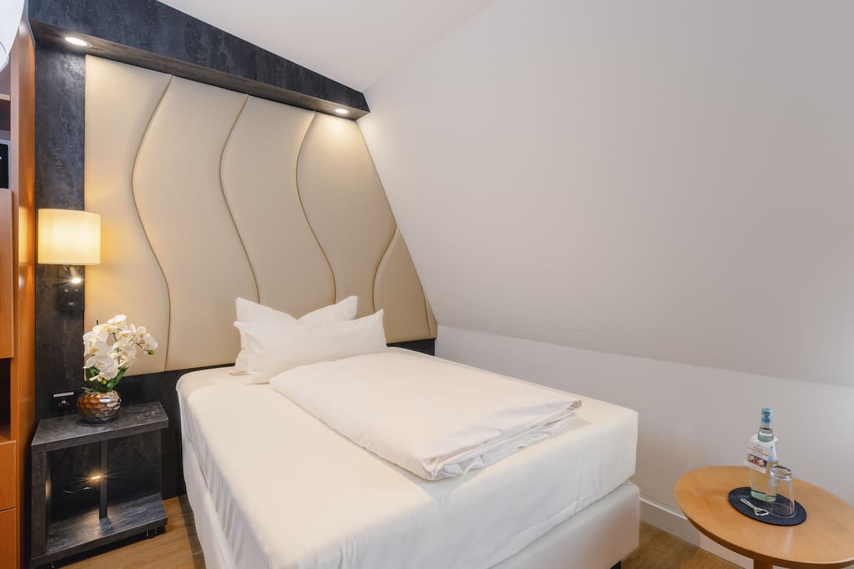 Cama bem arrumada com lençóis brancos, mesa de cabeceira com abajur e uma pequena mesa com garrafa e copo.