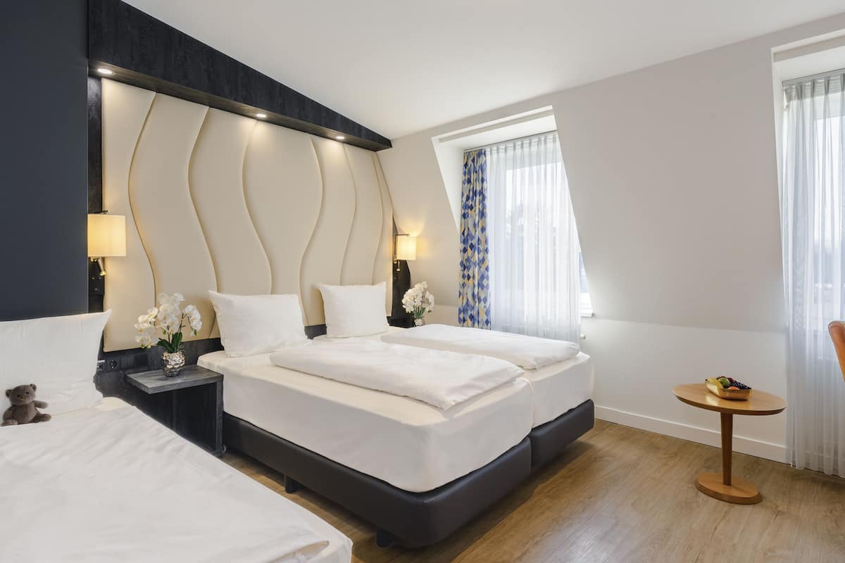 Quarto de hotel com uma cama grande, duas mesas de cabeceira com luminárias, uma mesinha redonda, uma cadeira e uma janela com cortinas.