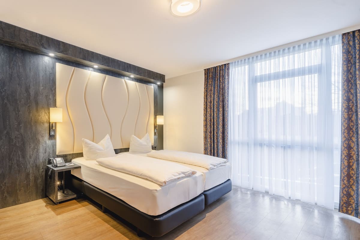Quarto de hotel moderno com uma cama grande, mesa de cabeceira com abajur e vista da cidade através de cortinas translúcidas.