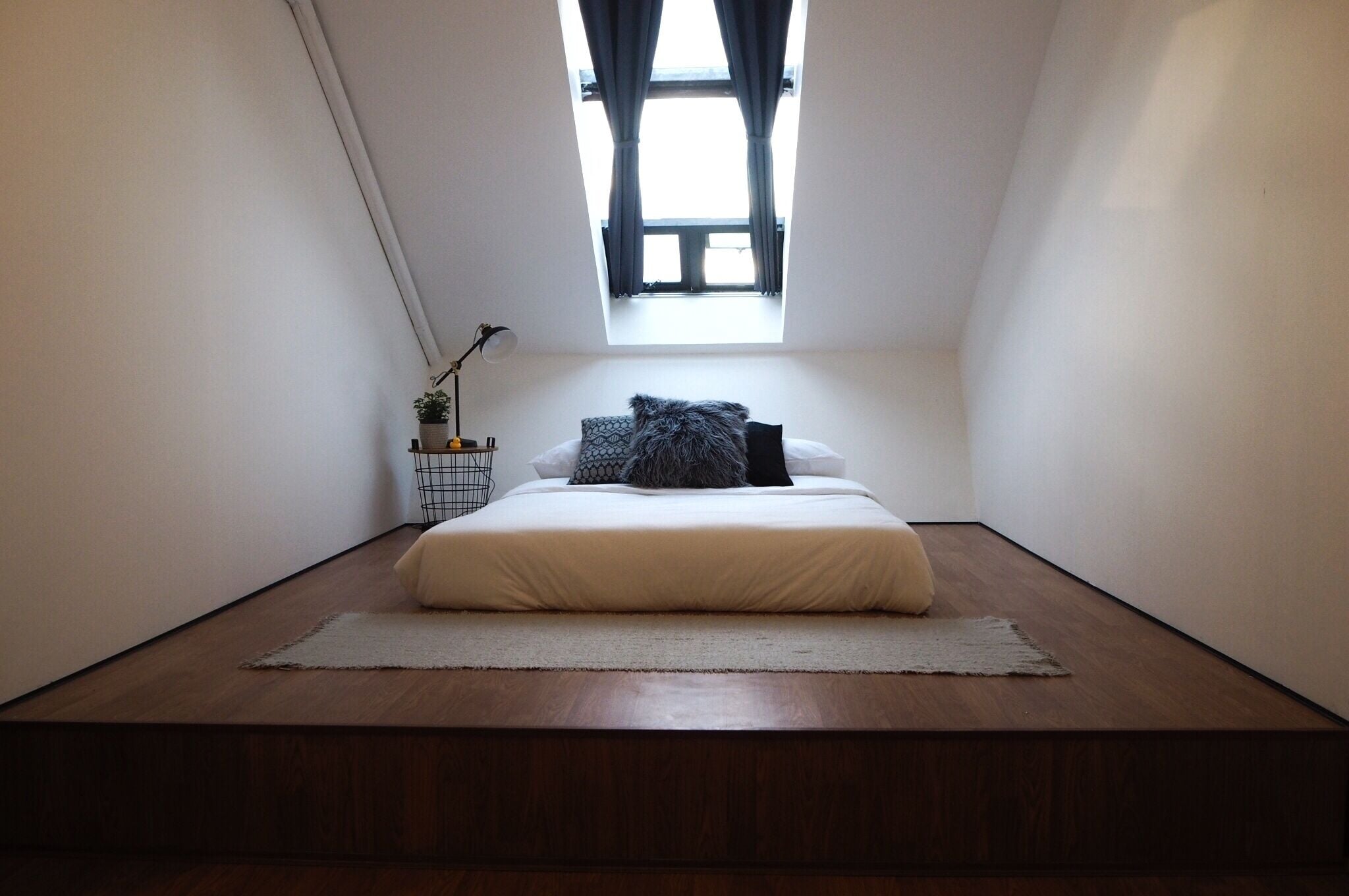 Quarto pequeno e minimalista com uma cama de solteiro, um claraboia e uma pequena mesa de cabeceira com uma planta.