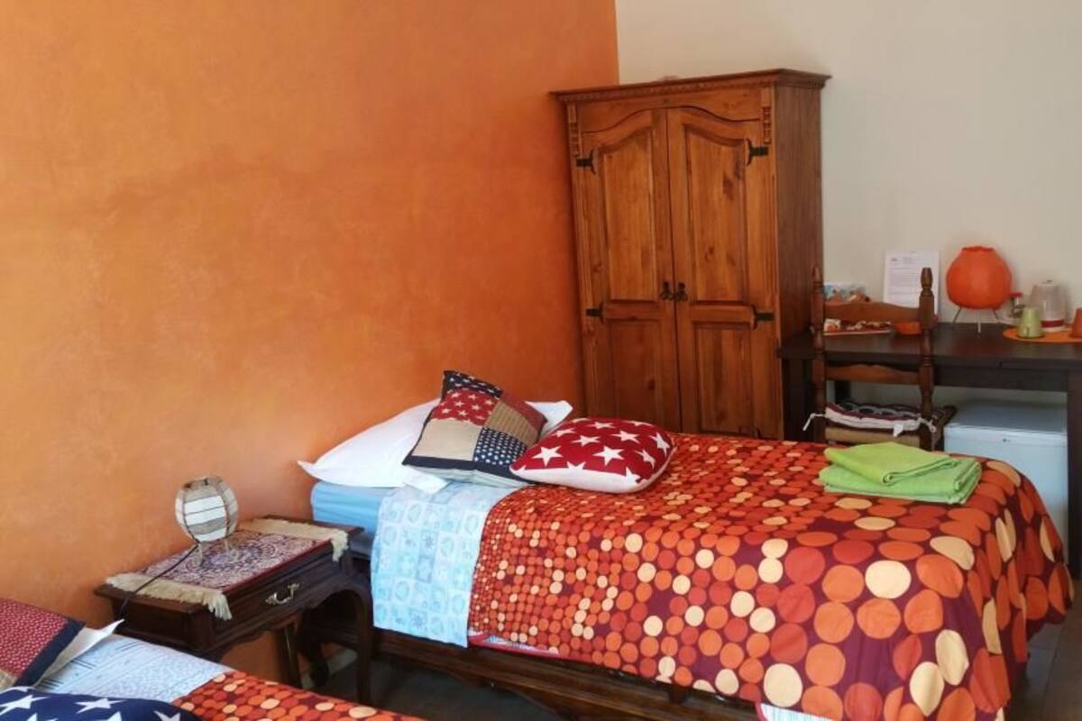 Quarto com cama, guarda-roupa de madeira, escrivaninha e uma mesinha de cabeceira com abajur.