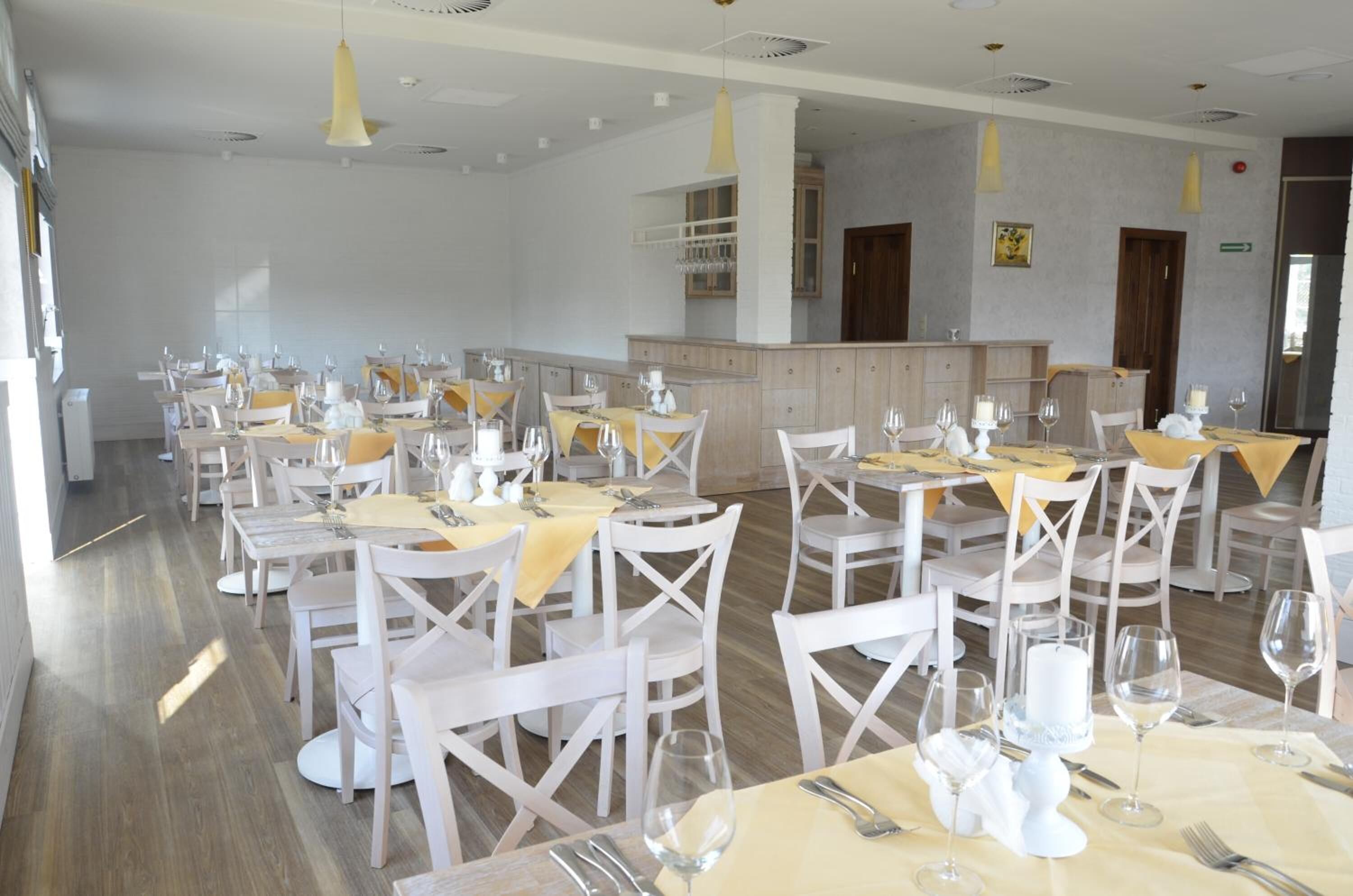 Uma sala de jantar com cadeiras e mesas brancas, toalhas de mesa amarelas e luminárias pendentes.