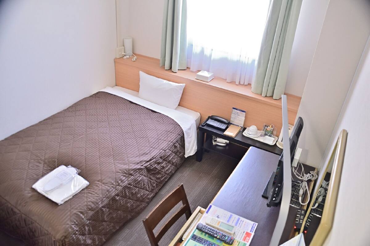 Quarto de hotel com cama, escrivaninha com computador, cadeira e janela com cortinas.