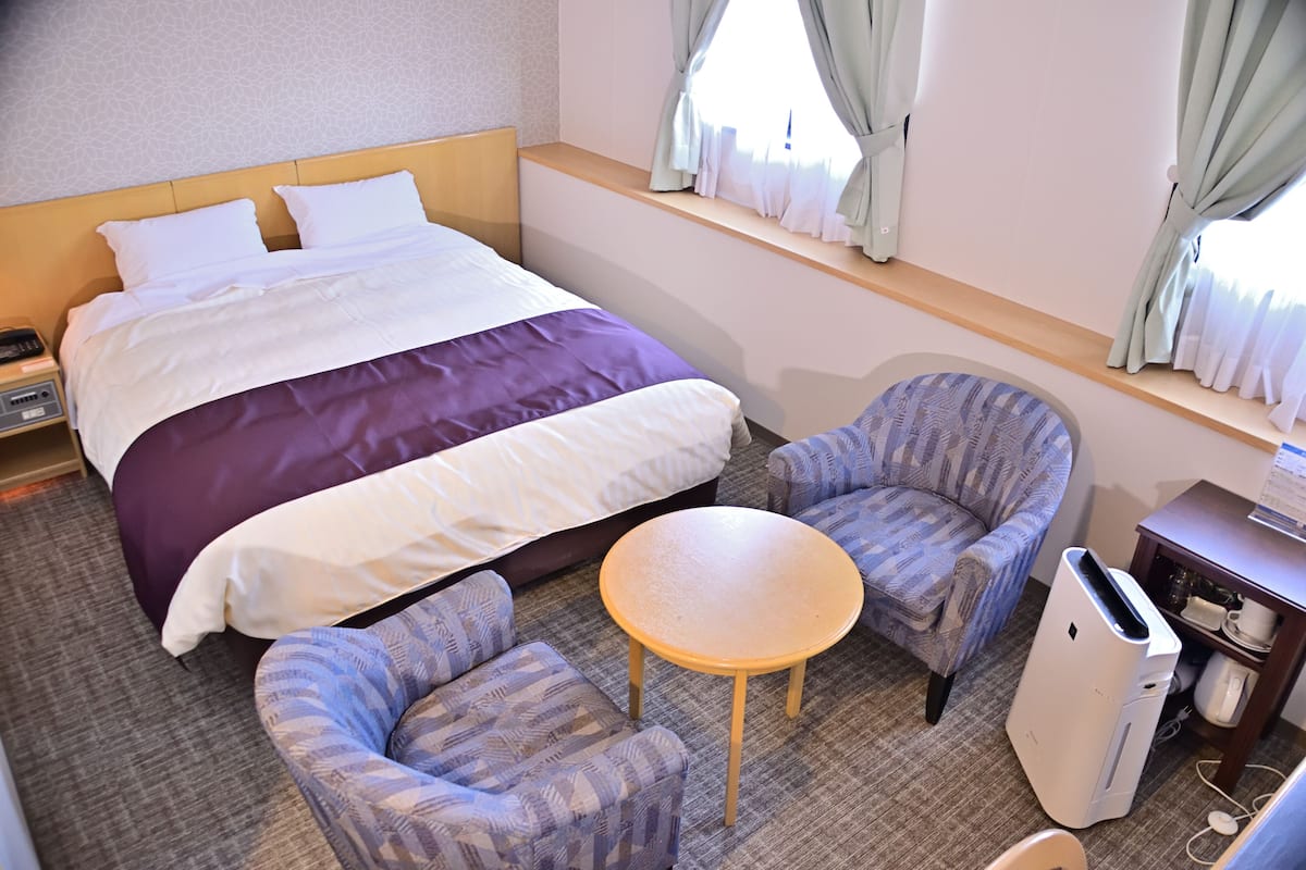 Quarto de hotel com uma cama, duas poltronas, uma mesa redonda pequena, um aquecedor e uma janela com cortinas.