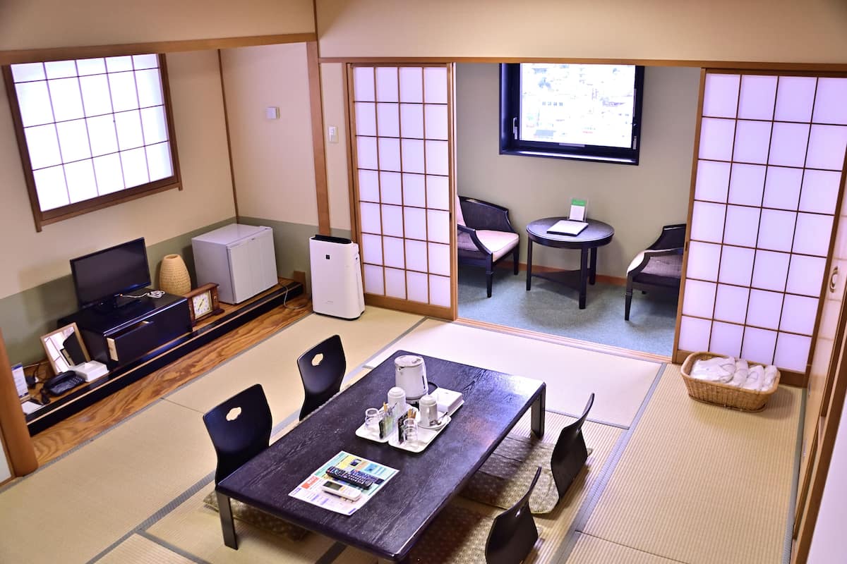 Quarto japonês tradicional com chão de tatame, mesa de tatame, cadeiras pretas, televisão, frigorífico e uma porta deslizante que dá para outro espaço.