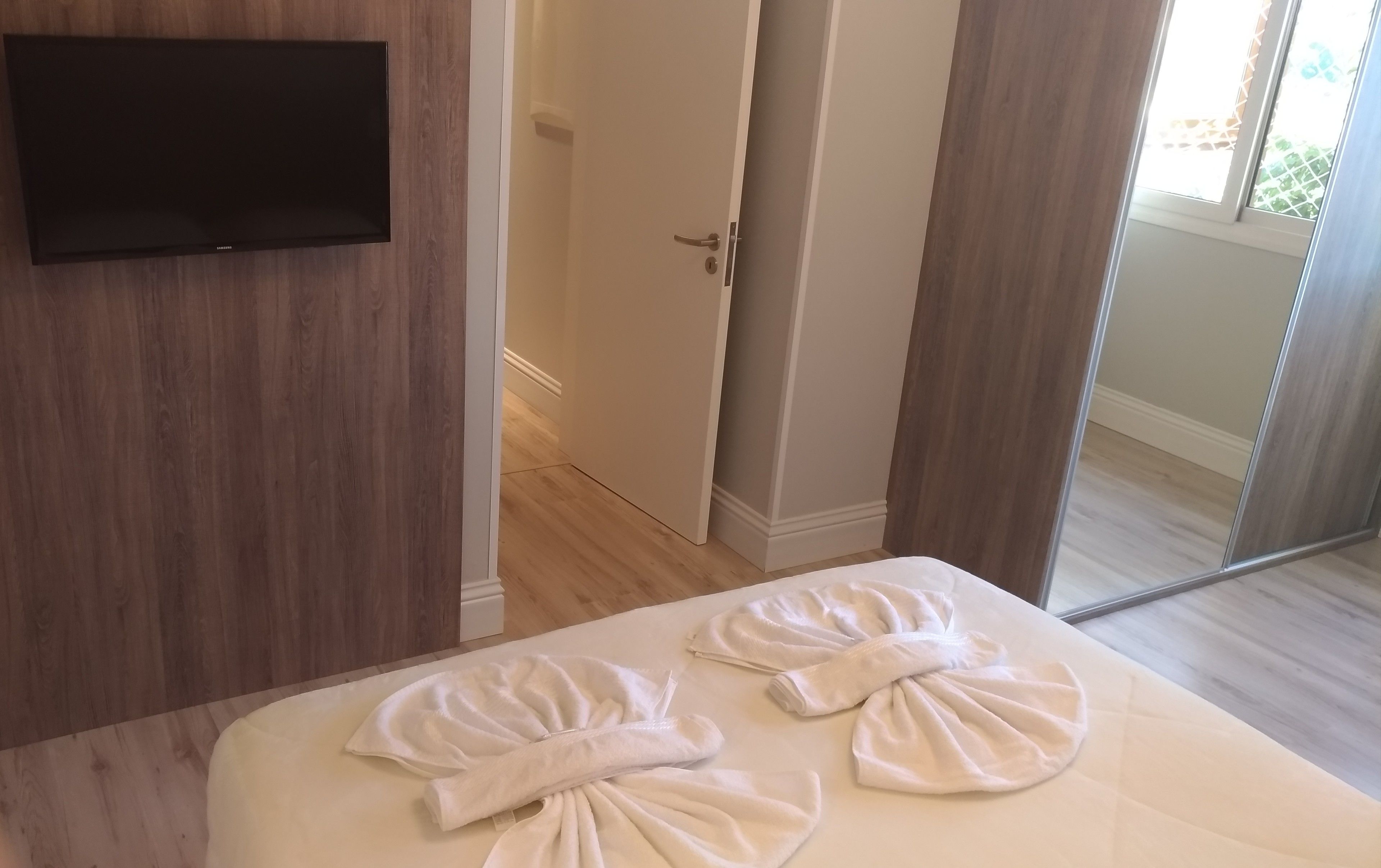Quarto de hotel com televisão de tela plana na parede, porta que dá para outro cômodo e cama com toalhas dobradas.