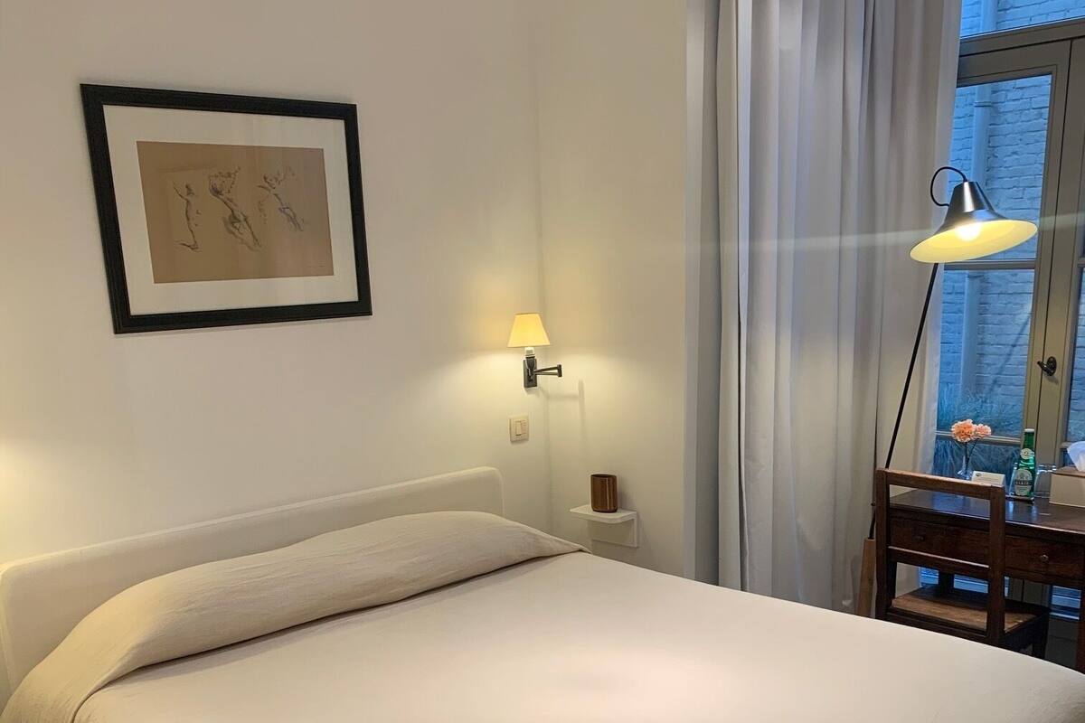 Cama bem arrumada com colcha branca, luminária fixada na parede e quadro emoldurado na parede.