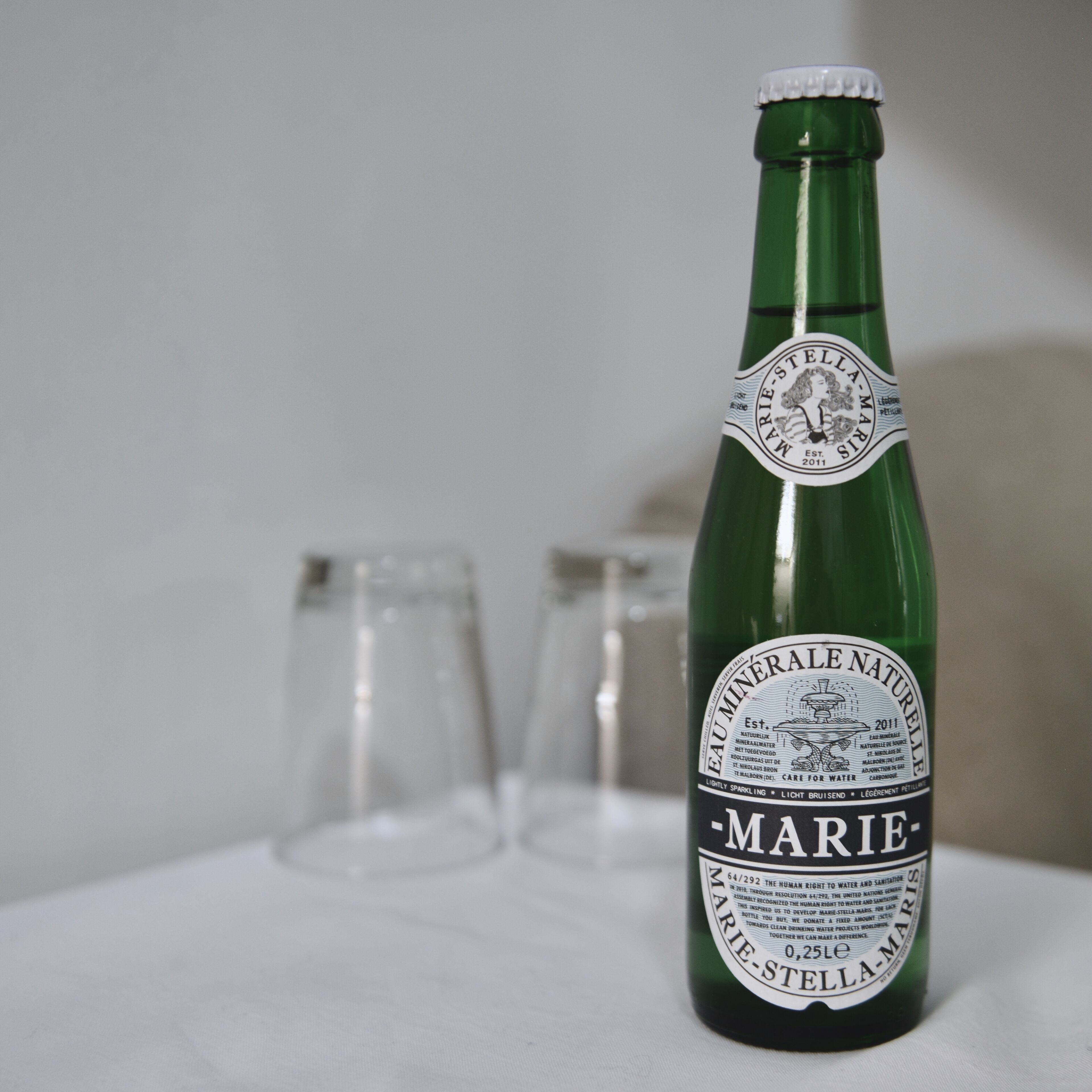 Uma garrafa de cerveja Marie-Stella-Mars com rótulo, colocada sobre uma mesa com dois copos vazios.