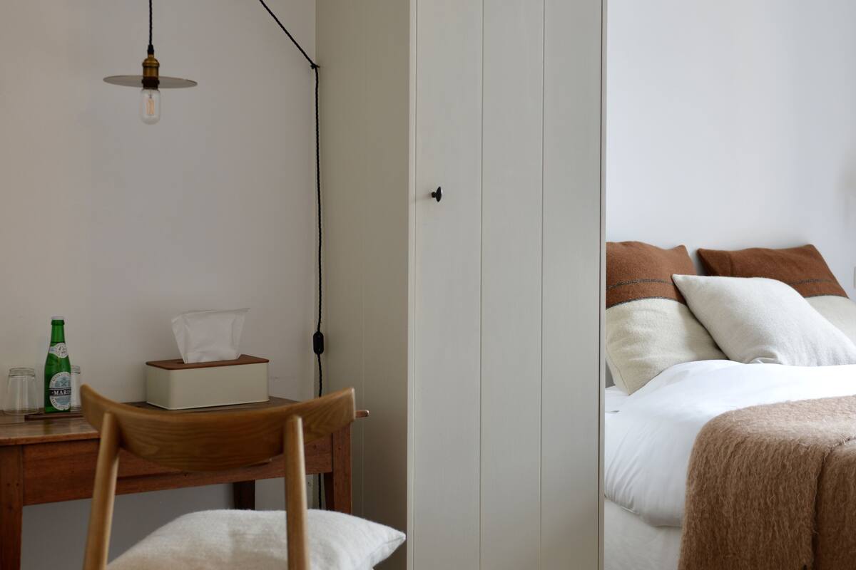 Quarto com cama, uma cadeira de madeira, escrivaninha com garrafa e guarda-roupa.